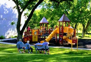Parquinho infantil com escorregadores e estruturas de escalada, cercado por árvores e um espaço com bancos.