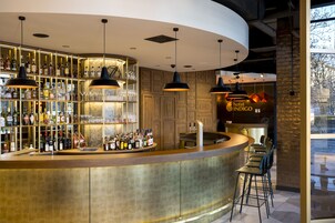 Uma área de bar com balcão curvo, prateleiras com garrafas de bebidas, luminárias pendentes e vista para o exterior através de uma grande janela.