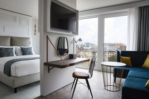 Quarto de hotel moderno com cama, escrivaninha, poltrona, TV e sofá.