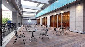 Uma área de café ao ar livre moderna, com mesas, cadeiras, guarda-corpo de vidro e vista para a paisagem urbana.