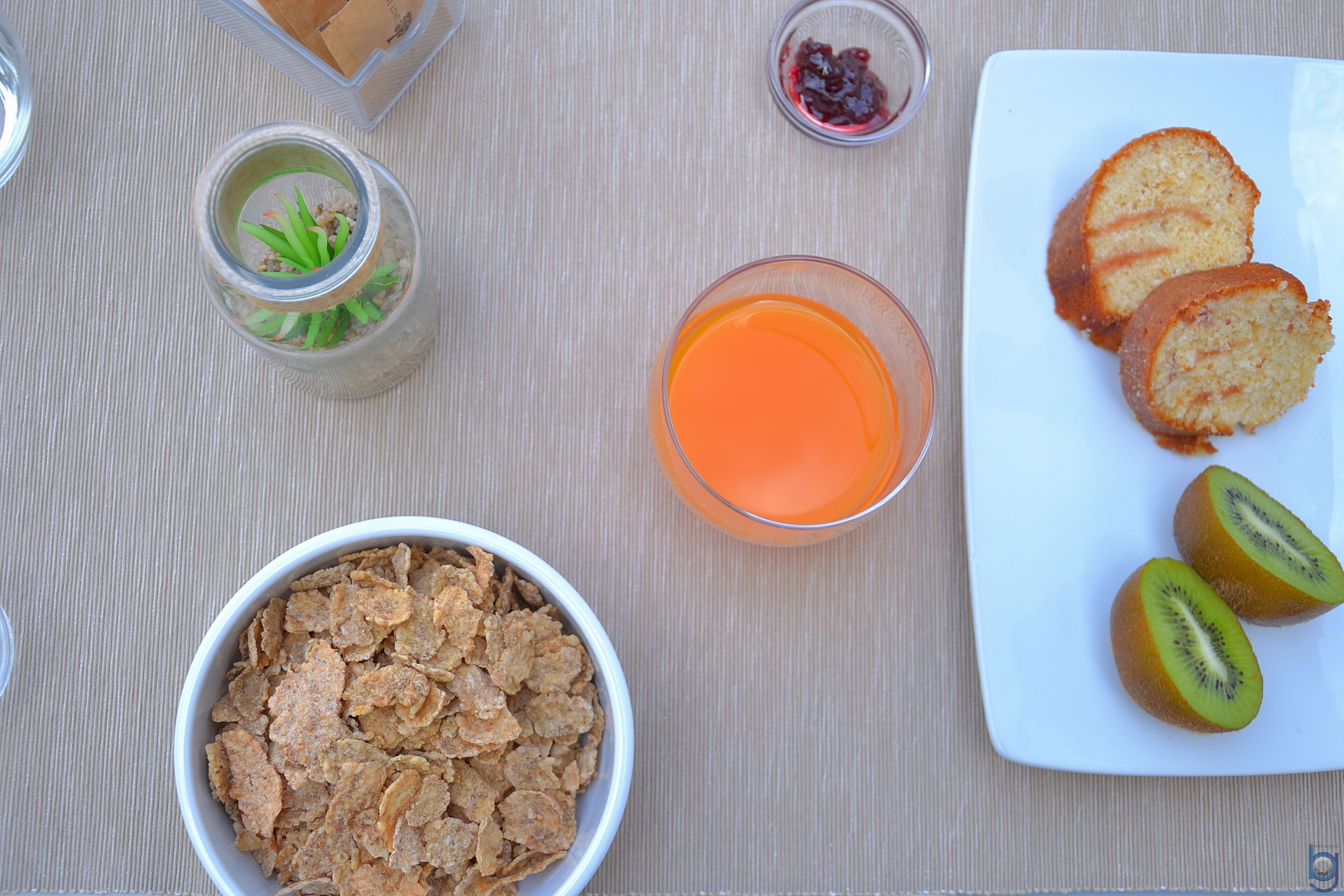 Mesa de café da manhã com cereais, fatias de pão, kiwi, copo de suco de laranja e um pequeno pote de geleia.