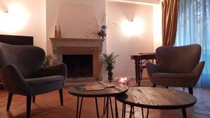Uma sala de estar com lareira, dois poltronas, uma mesa redonda de madeira e um vaso com planta.