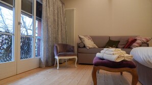 Um quarto com piso de madeira, um sofá cinza com almofadas, uma poltrona pequena, um banco de madeira com toalhas e uma porta de vidro deslizante com cortinas.
