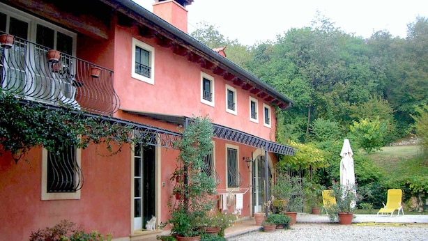 Um edifício de dois andares com fachada vermelha, uma varanda com grades de metal e um pátio de cascalho com plantas em vasos e móveis de exterior.