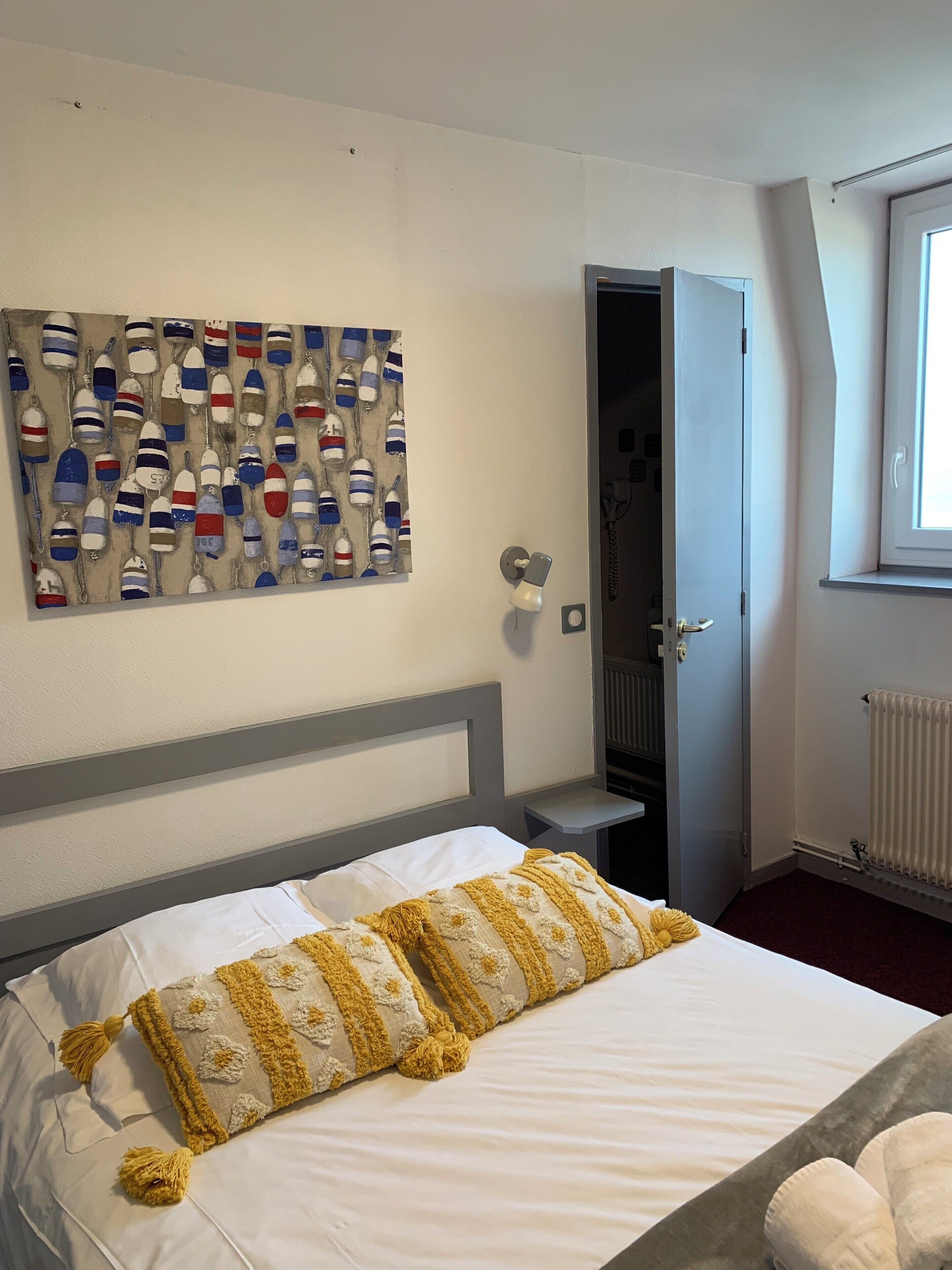 Quarto de hotel com uma cama, uma obra de arte na parede representando bóias, um radiador e uma porta que dá para o banheiro.