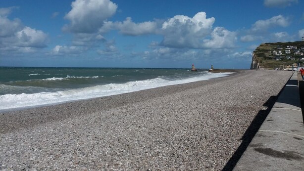 Uma praia de seixos com um farol ao longe, um cais que se estende para o mar e casas em um penhasco.