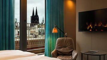Quarto de hotel com cama, poltrona, mesa pequena, televisão e vista para a paisagem urbana com uma catedral.