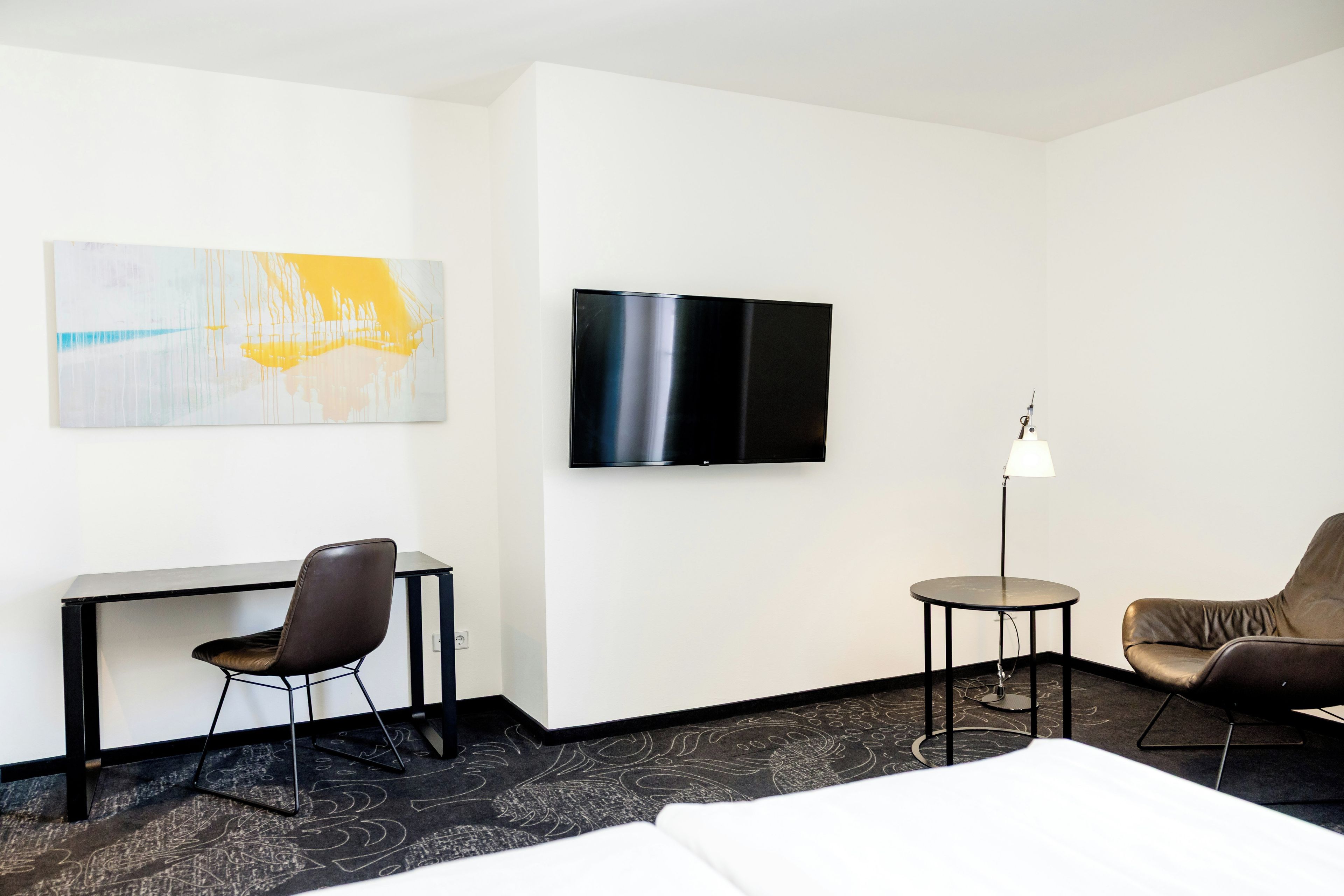 Quarto de hotel com cama, escrivaninha, cadeira, televisão e luminária.