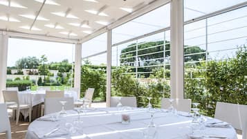 Uma área de refeições ao ar livre coberta, com mesas redondas preparadas para uma refeição, cercada por grandes janelas que oferecem vista para um jardim exuberante.