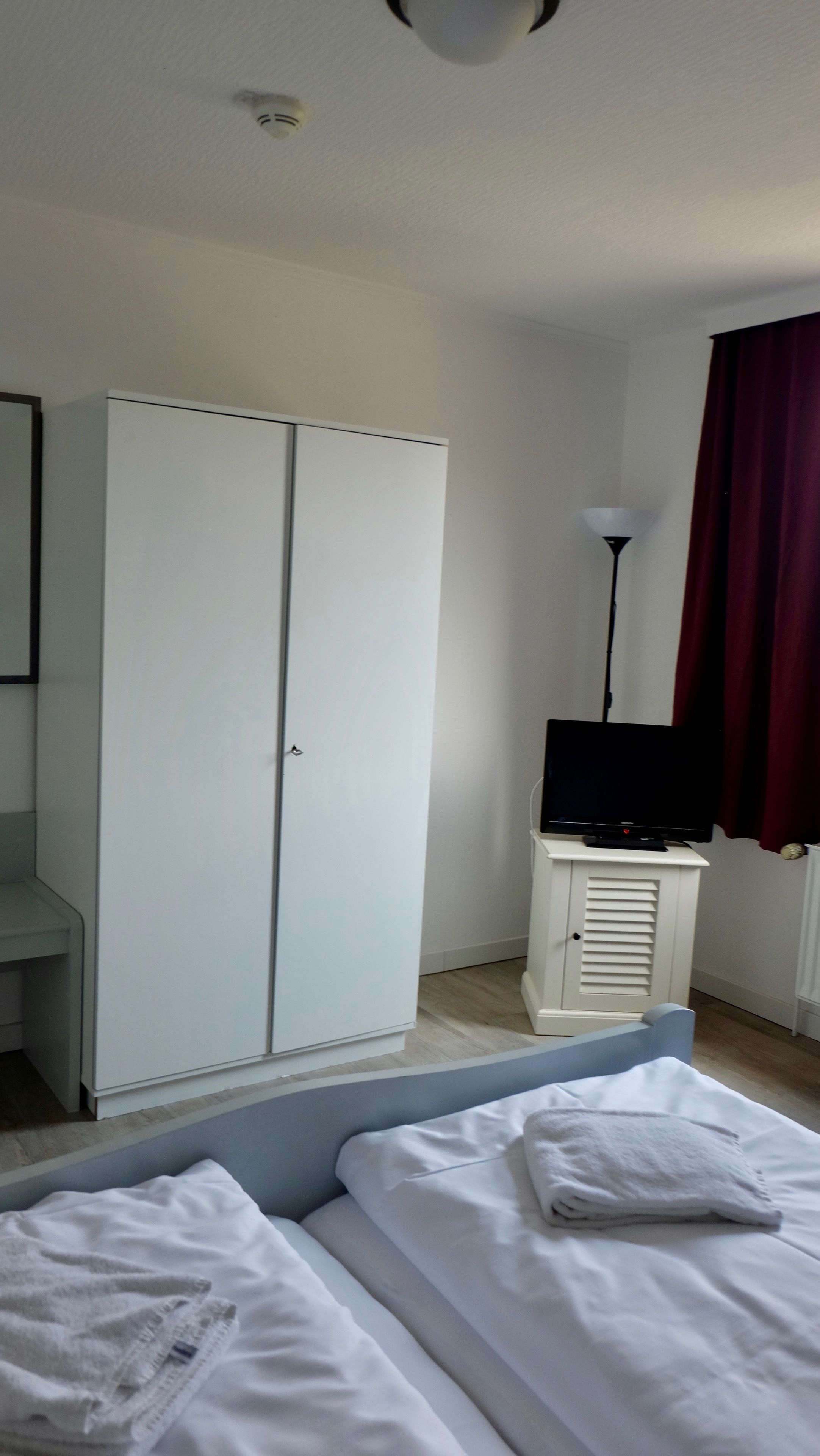 Quarto com cama, televisão sobre suporte, guarda-roupa e luminária no teto.
