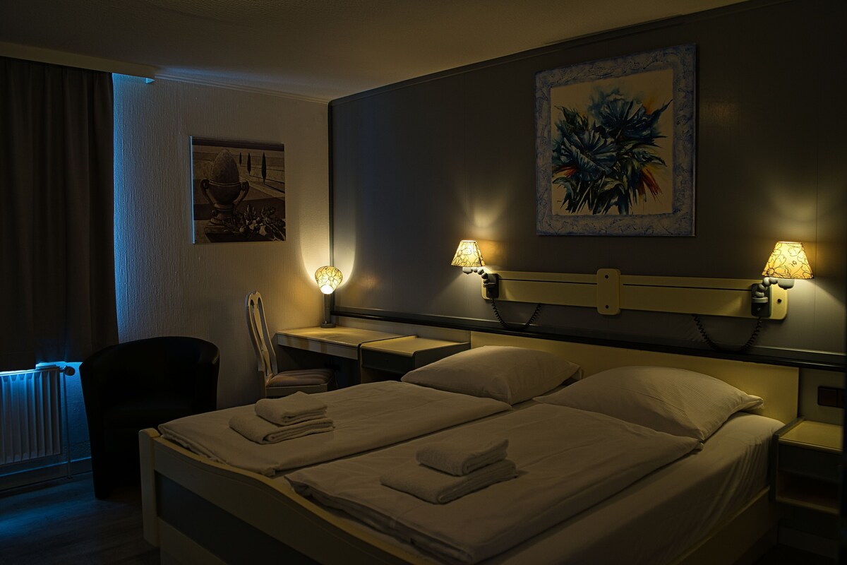 Quarto de hotel com duas camas, uma mesa de cabeceira, uma escrivaninha, uma cadeira e um radiador.