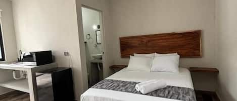 Quarto de hotel com cama de madeira, escrivaninha com computador e banheiro visível por uma porta aberta.