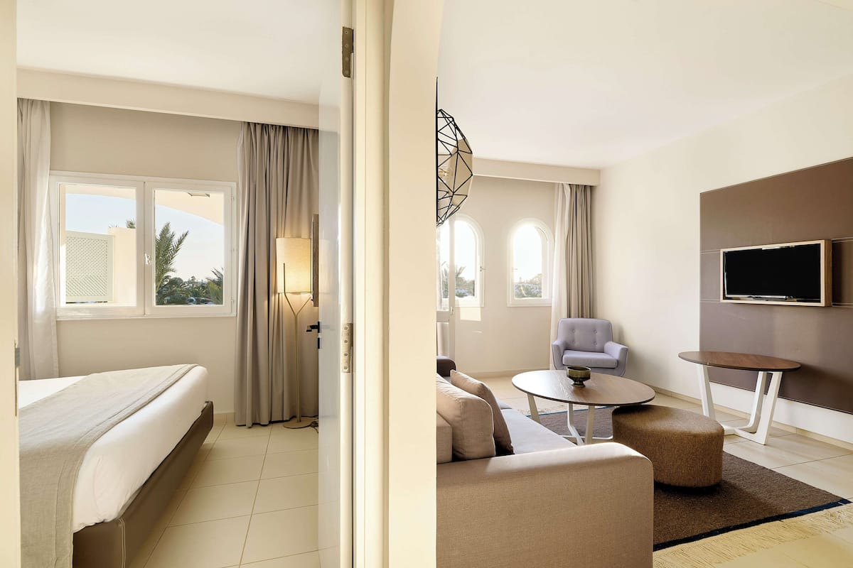 Quarto de hotel com cama, sofá, mesa pequena, televisão e janela com cortinas.