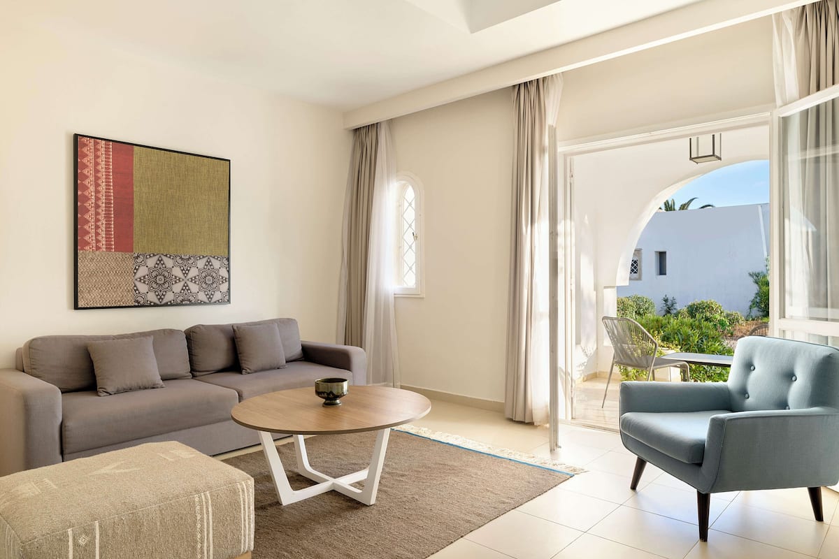 Uma sala de estar moderna com sofá, poltrona e mesa de centro, uma janela grande com cortinas e vista para o verde lá fora.