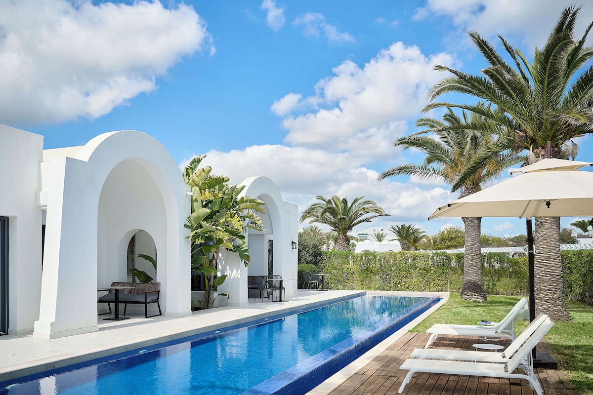 Uma área de piscina com piscina azul cristalina, arcos brancos, espreguiçadeiras e palmeiras.