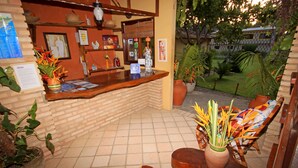 Uma área de recepção com balcão de madeira, plantas em vasos e uma cadeira com almofada florida.
