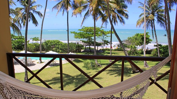 Uma rede numa varanda com vista para uma praia, palmeiras e uma visão clara do oceano.