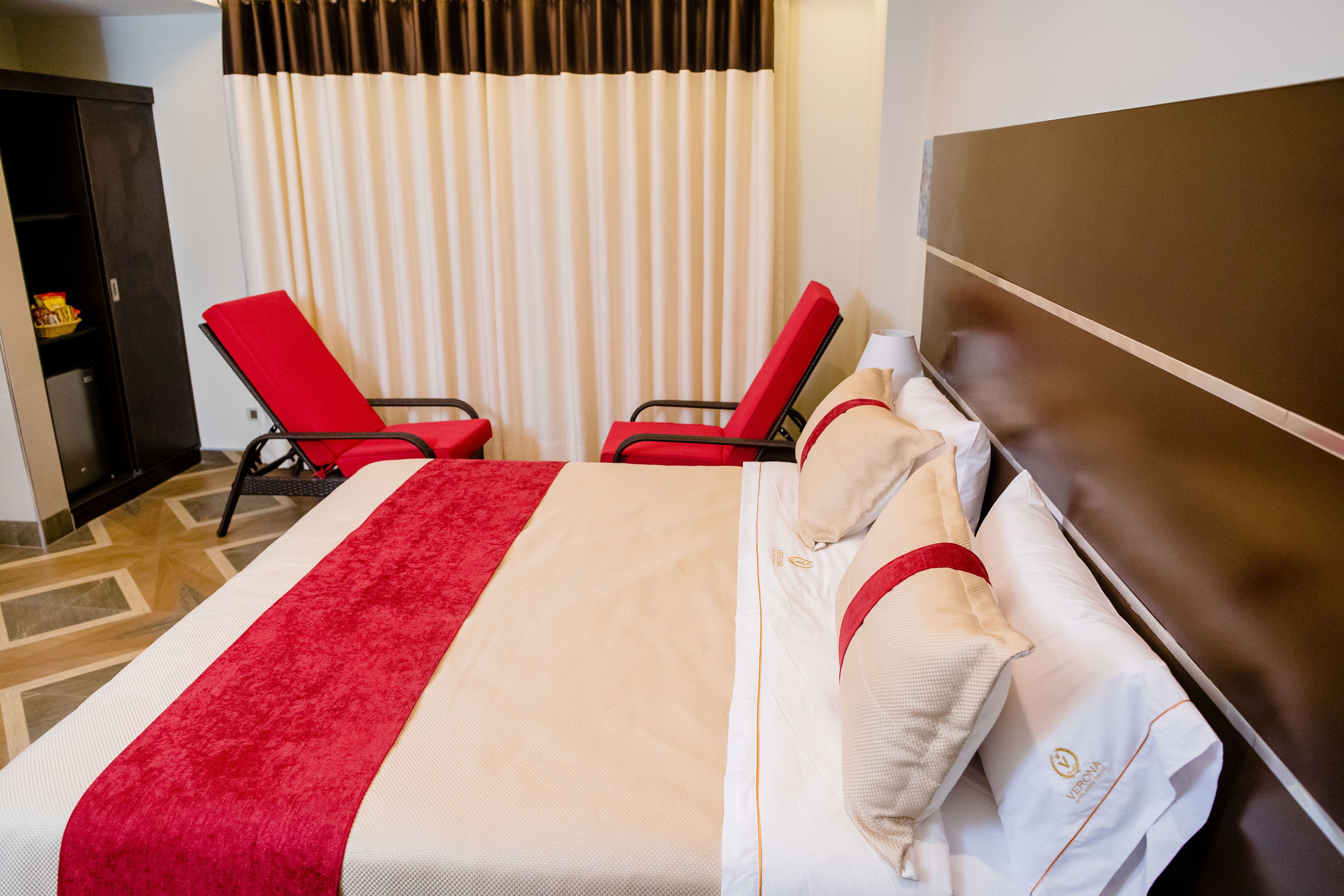 Quarto de hotel com uma cama, duas cadeiras vermelhas, um cobreleite vermelho e branco e uma janela grande com cortinas.