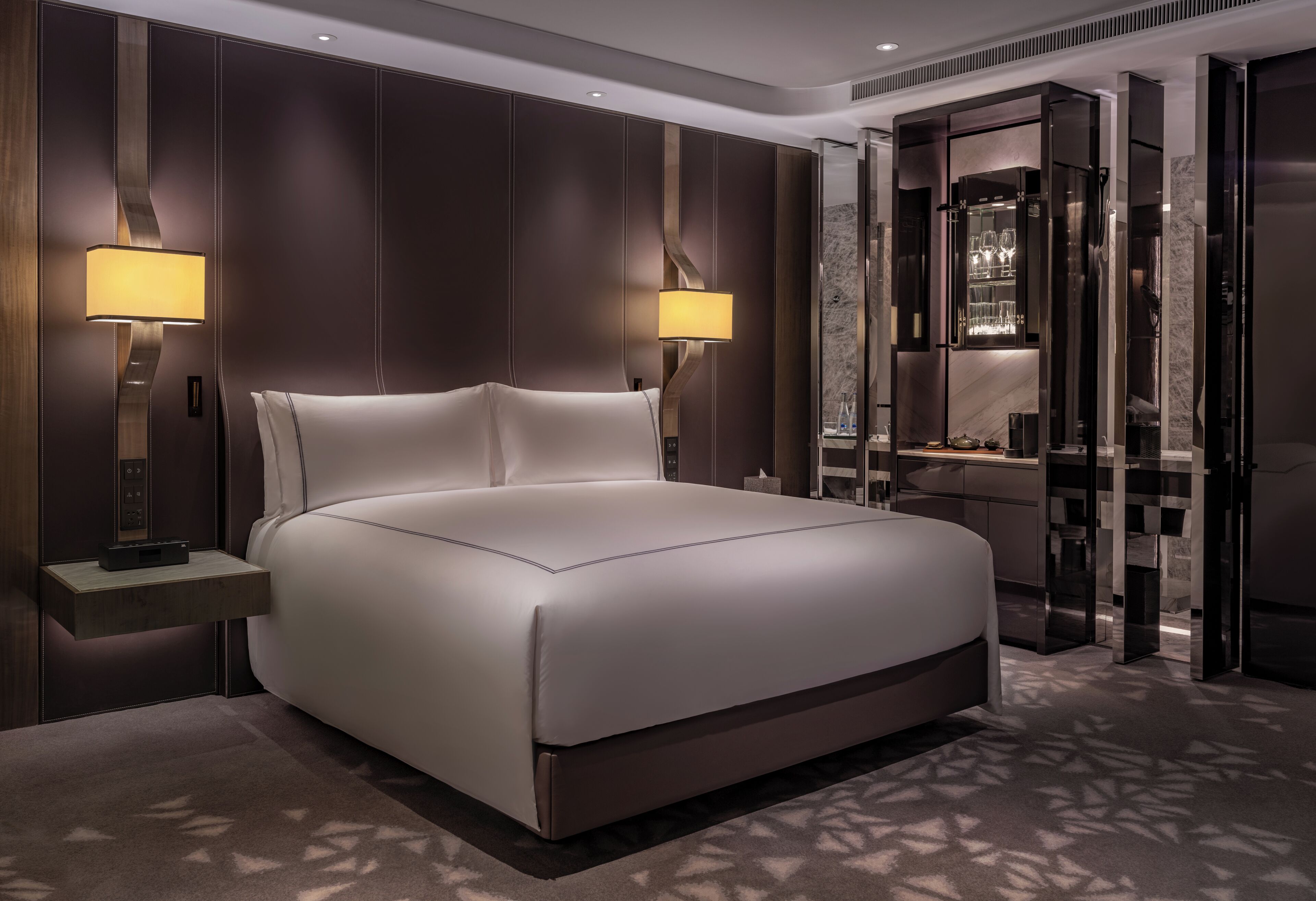 Quarto de hotel moderno com uma cama grande, duas luminárias de cabeceira, televisão integrada e minibar.