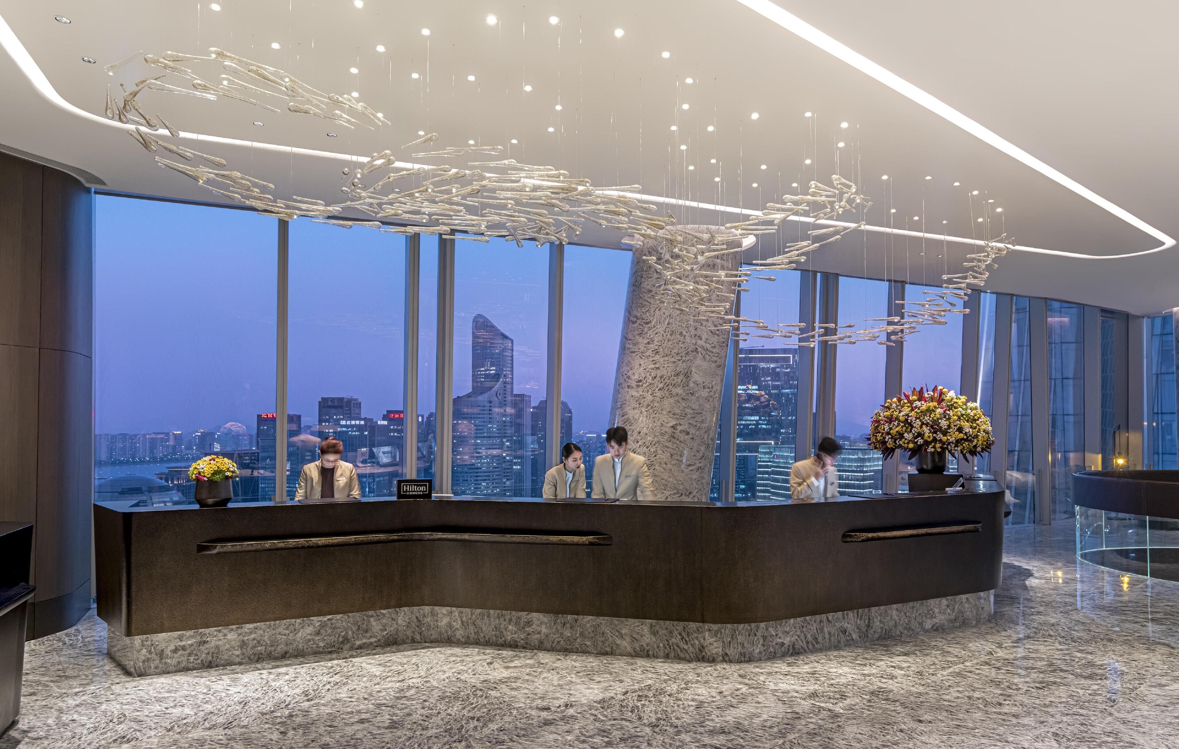 Uma área de recepção de hotel moderna, com uma grande janela que oferece vista para a cidade, um balcão de recepção de mármore e uma luminária de teto singular.