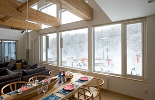 Uma sala de jantar com mesa de madeira posta para quatro pessoas, grandes janelas com vista para a neve e um espaço de estar acolhedor com sofá e cadeiras.