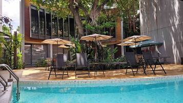 Uma piscina com espreguiçadeiras e guarda-sóis, cercada por plantas e um edifício com grandes janelas.