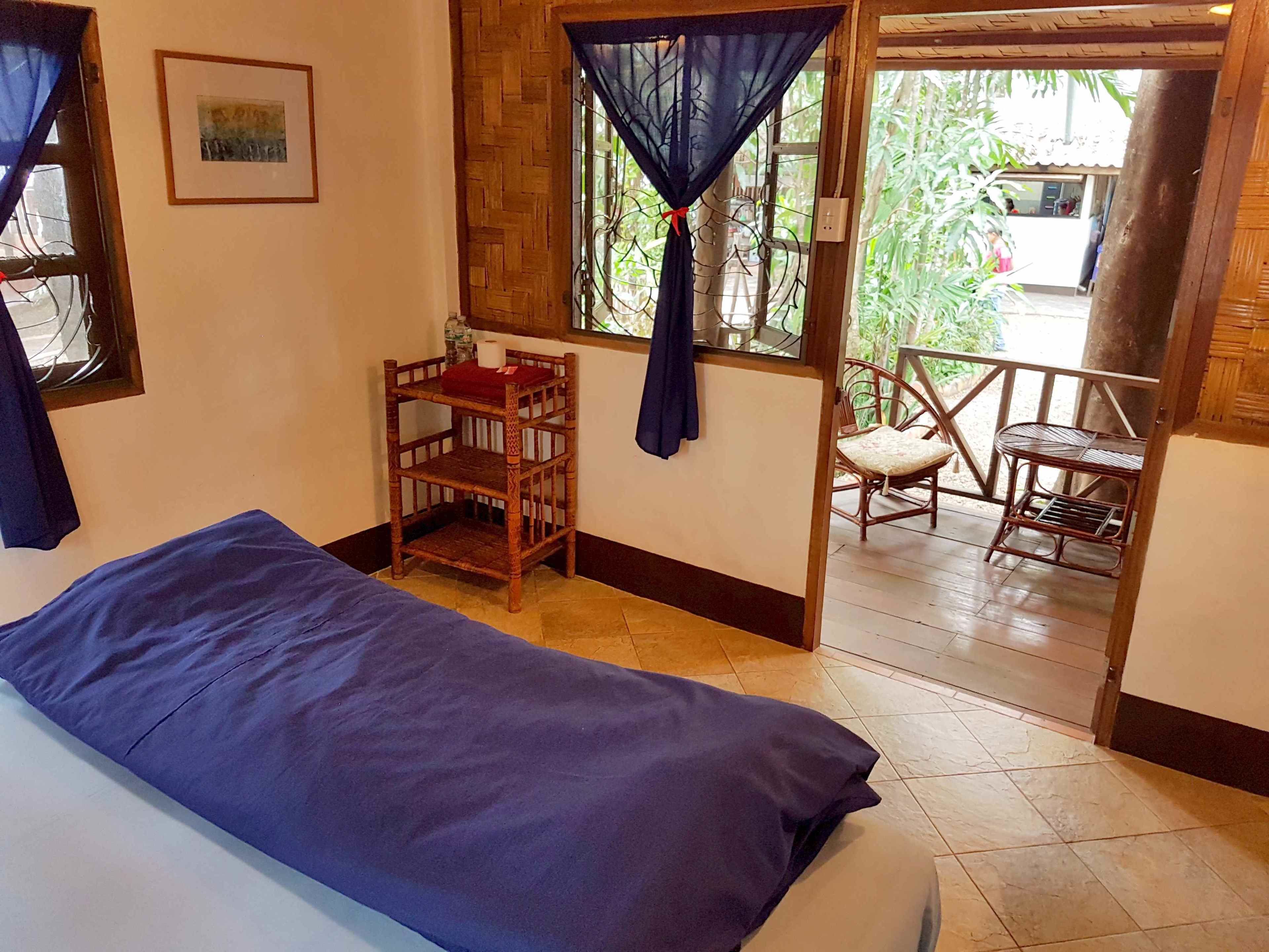 Quarto com uma cama, uma pequena mesa de madeira com um tecido vermelho, uma cadeira e uma janela com cortina azul.