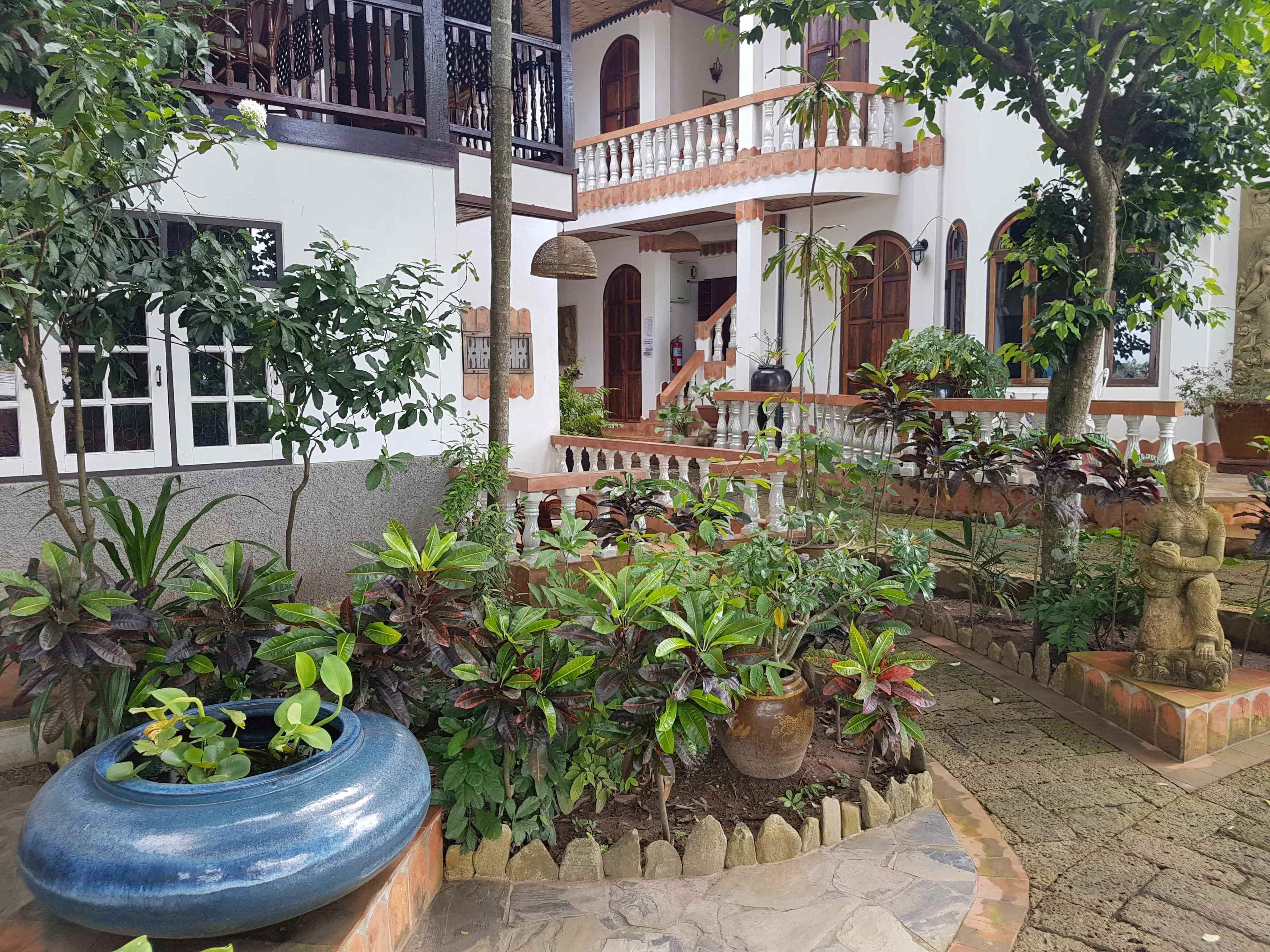 Um pátio com plantas em vasos, um caminho de pedra e um edifício com varanda e escadaria.