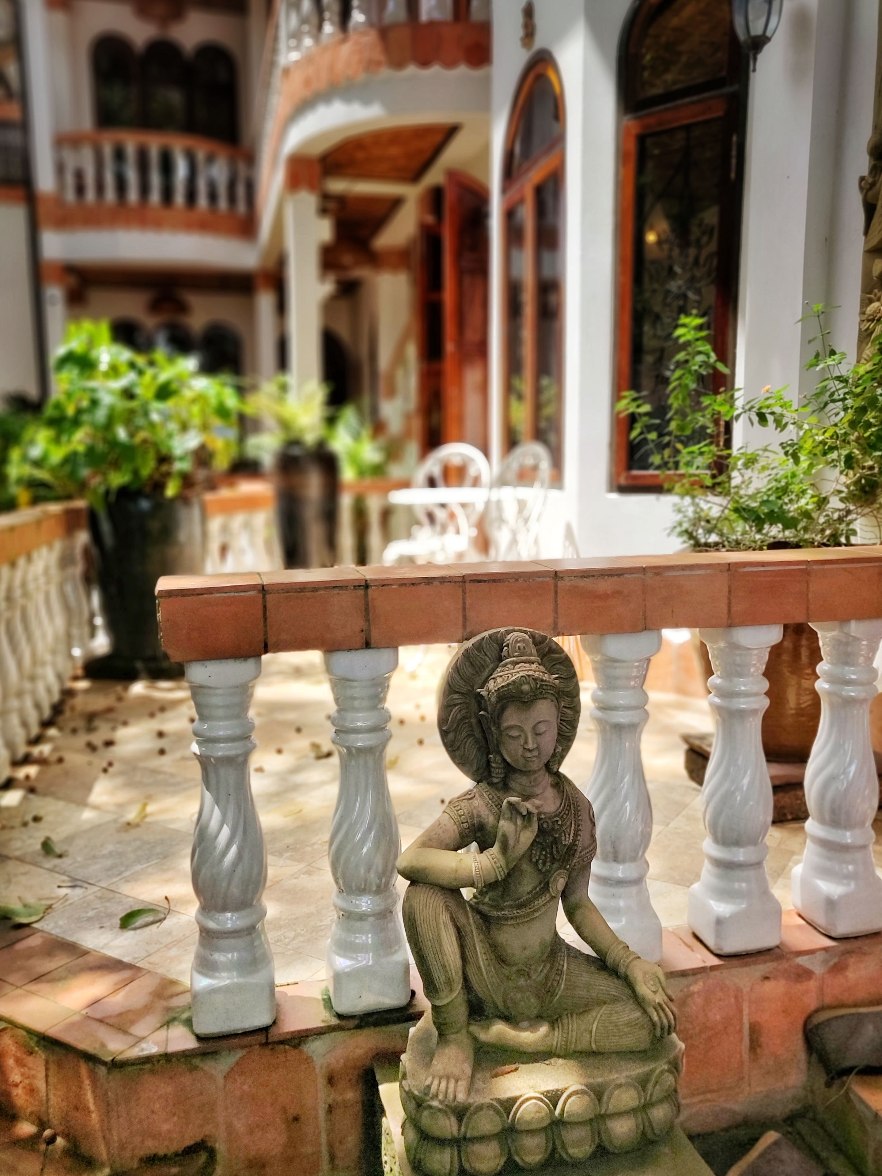 Uma estátua de uma figura sentada sob um parapeito branco, com piso de azulejos e plantas em vasos ao fundo.
