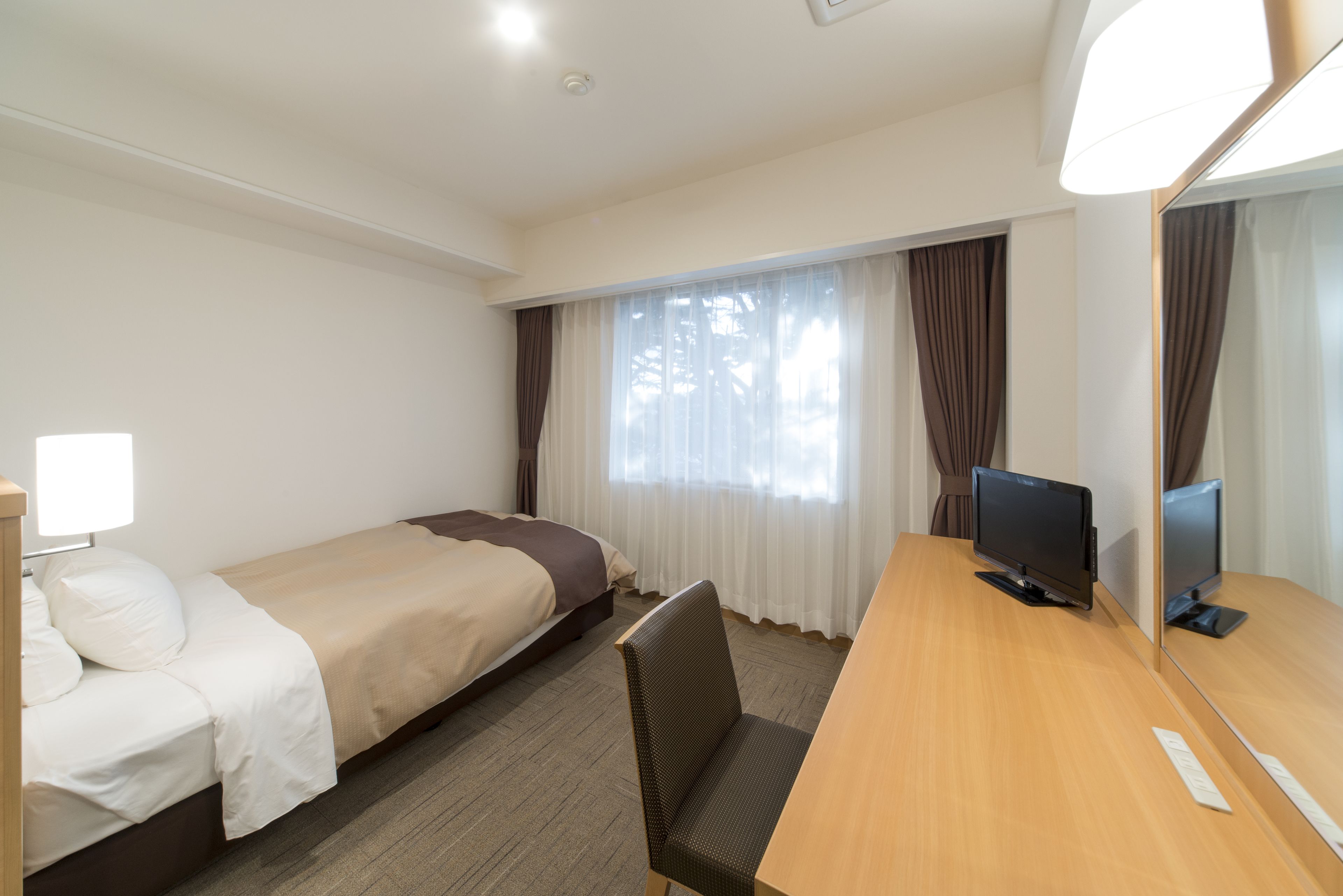 Quarto de hotel com uma cama de solteiro, uma mesa com computador, uma cadeira e uma janela grande com cortinas.