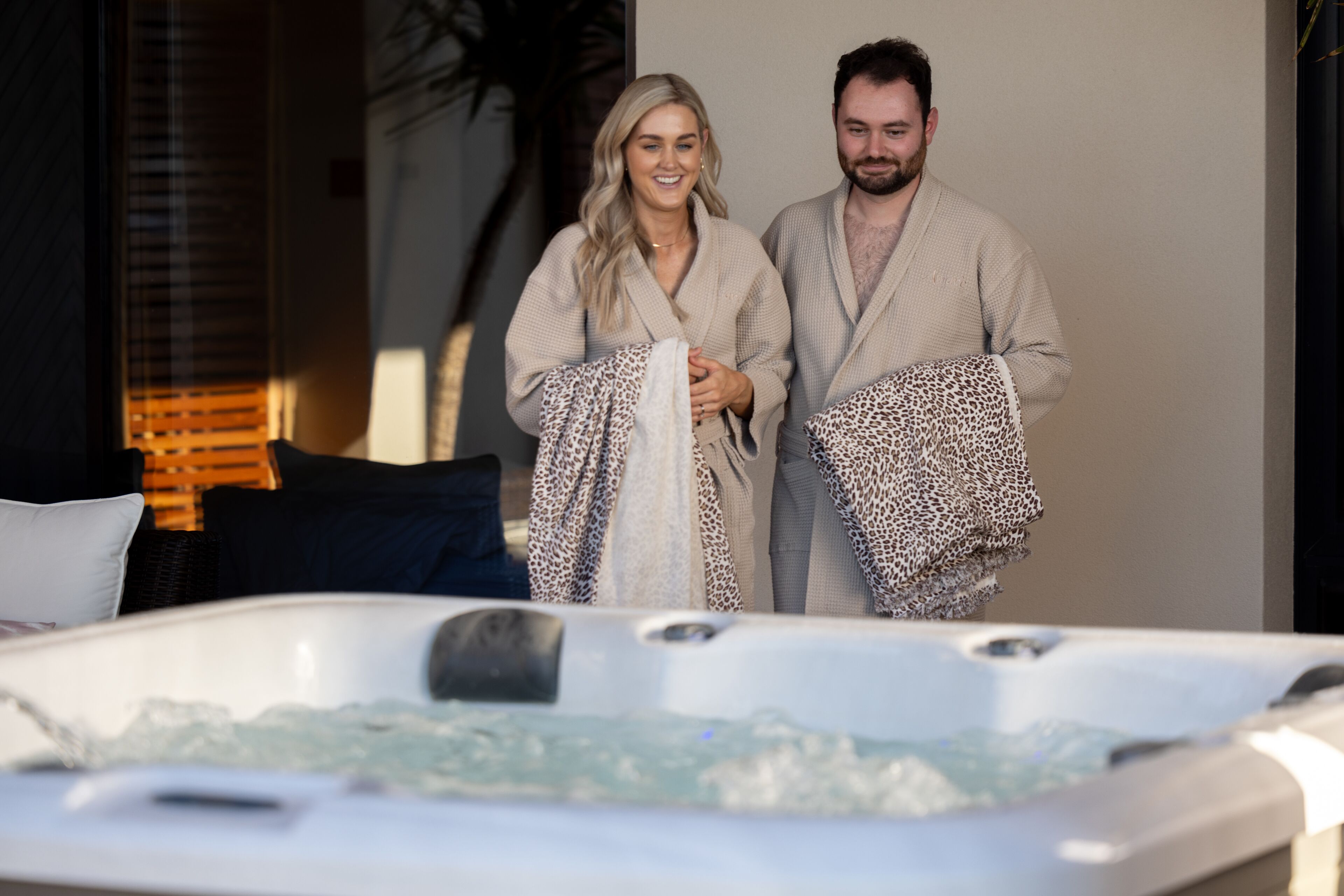 Um casal em roupões segurando toalhas, em frente a uma banheira de hidromassagem.