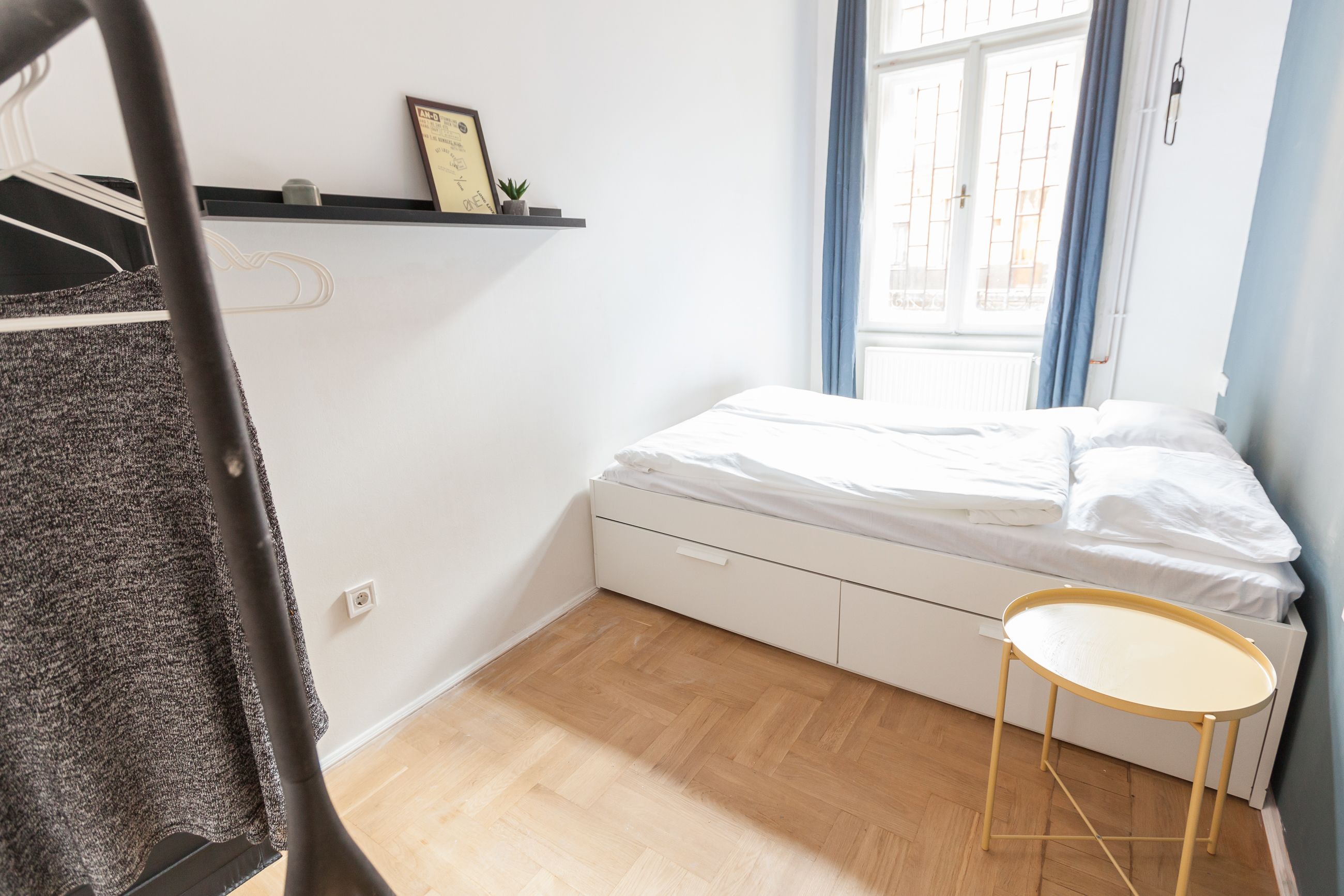 Quarto com cama, uma mesinha, uma estante com moldura e janela com cortinas.