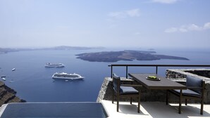 Terraço à beira-mar com mesa e cadeiras, de frente para um mar tranquilo e navios de cruzeiro.