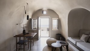 Uma sala de estar aconchegante com sofá, mesa de jantar com cadeiras e uma pequena mesa redonda com banquetas. Há duas janelas que proporcionam luz natural, e é visível uma vista do exterior pela porta aberta.