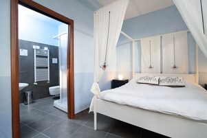 Quarto com cama, mesa de cabeceira, janela com cortinas, banheiro com vaso sanitário, pia e porta-toalhas.