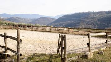 Um espaço ao ar livre cercado por uma cerca, com chão de terra, rodeado por colinas e uma aldeia distante.