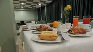 Disposição de mesa para o café da manhã com croissants, suco de laranja e café, em um ambiente com cadeiras e cortinas verdes.
