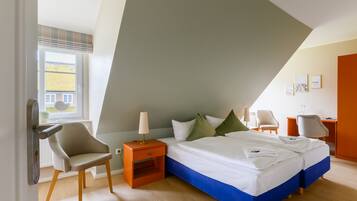 Quarto com teto inclinado, cama com roupa de cama branca e azul, duas cadeiras, mesa de cabeceira com abajur e janela com vista para um edifício.