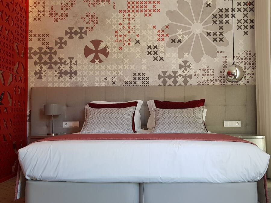 Quarto de hotel com cama, mesa de cabeceira, abajur e parede estampada.