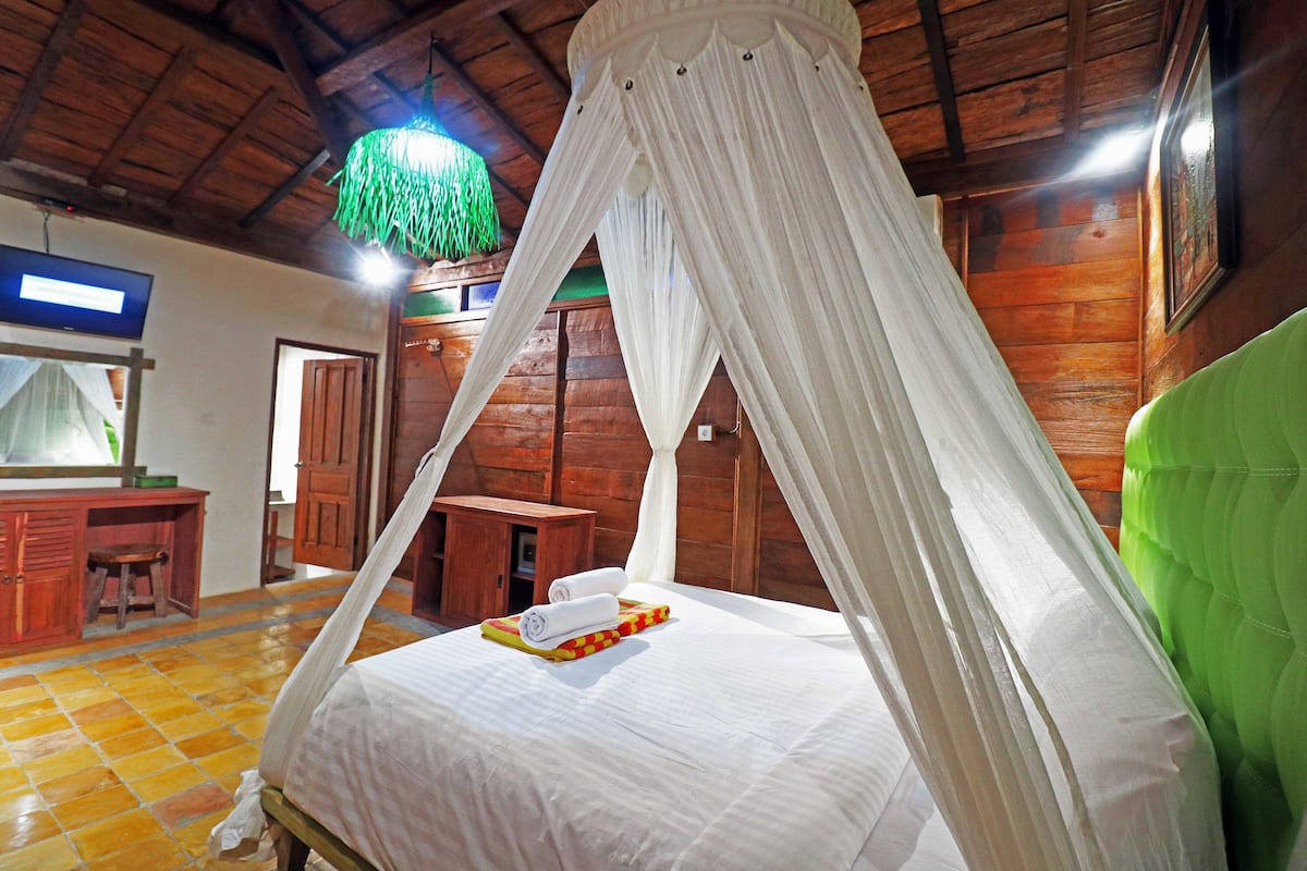 Quarto com cama sob um dossel branco, uma escrivaninha de madeira e uma televisão.