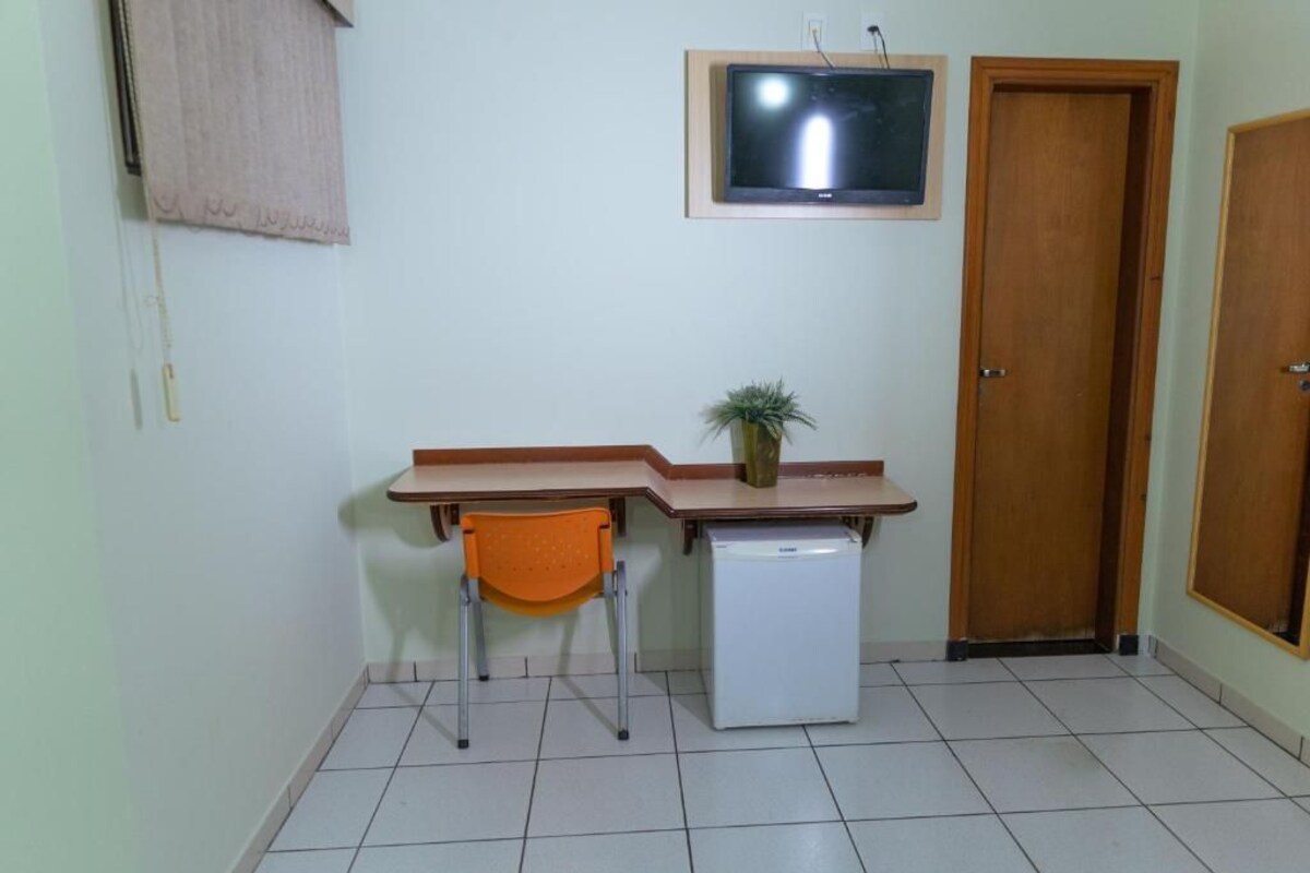 Quarto pequeno com escrivaninha, cadeira, televisão na parede, frigorífico e duas portas.