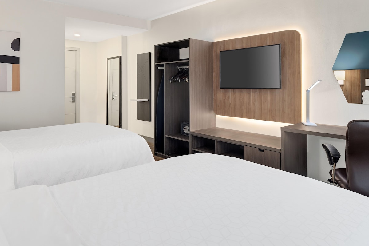 Quarto de hotel com duas camas, uma televisão na parede, uma escrivaninha com abajur e um guarda-roupa.