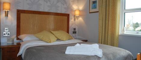 Quarto de hotel com uma cama, duas luminárias de cabeceira, uma mesa de cabeceira, uma janela com cortinas e uma parede com papel de parede estampado.
