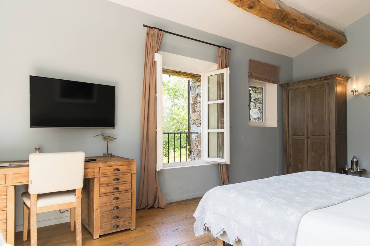 Quarto com cama, escrivaninha com cadeira, televisão, janela com cortinas e guarda-roupa.