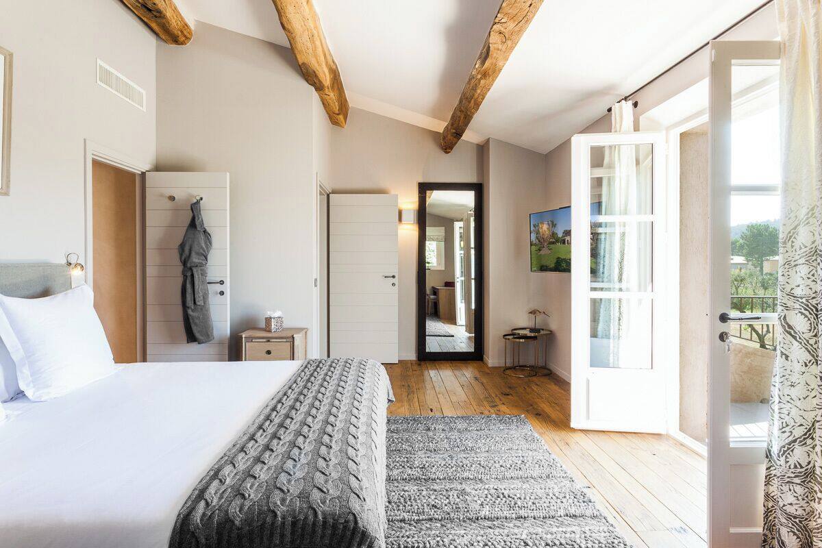 Quarto com cama grande, vigas de madeira, porta para uma varanda e vista para o exterior.