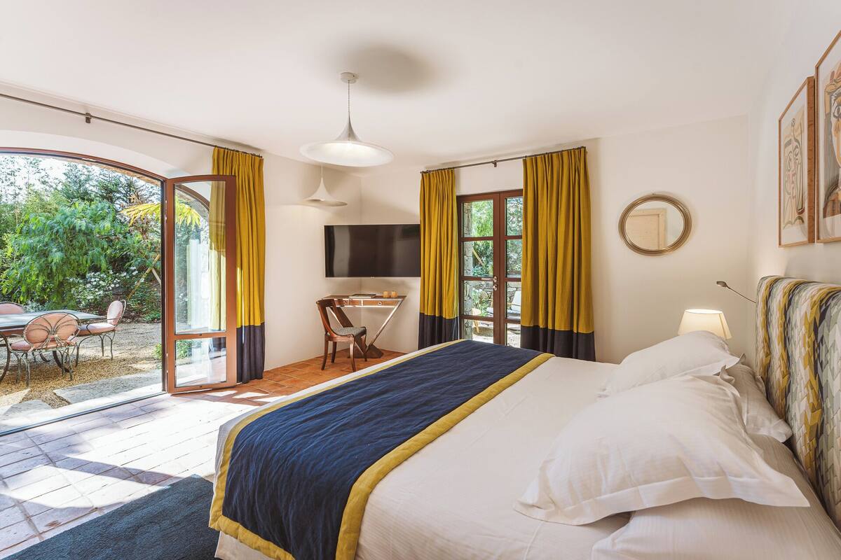 Quarto com cama, escrivaninha, cadeira, televisão e vista para o exterior.