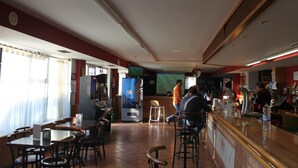 Um restaurante com mesas e cadeiras altas, área de bar e uma tela de televisão.