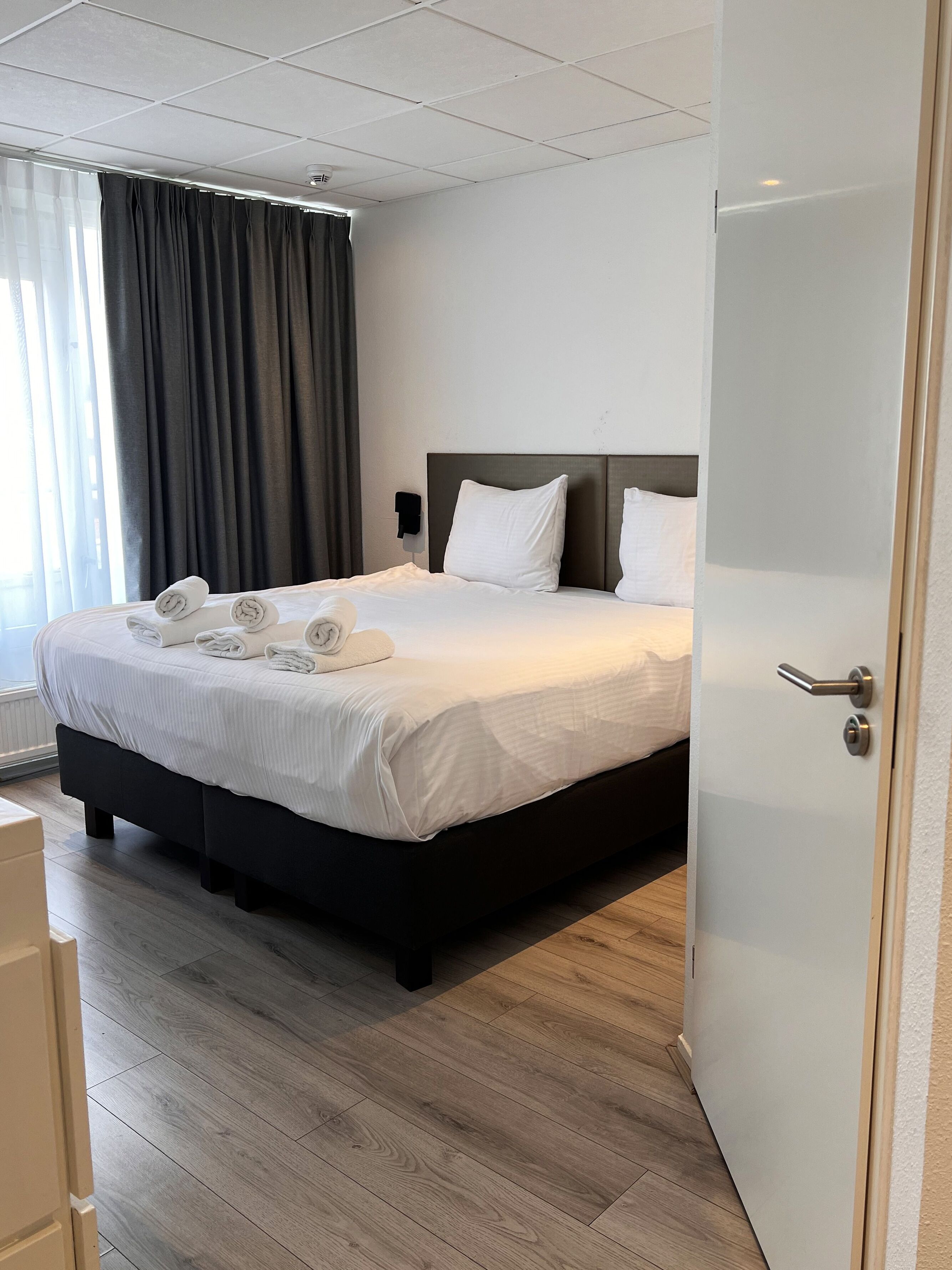 Quarto de hotel com cama bem arrumada, mesa de cabeceira, telefone fixado na parede e janela com cortinas.