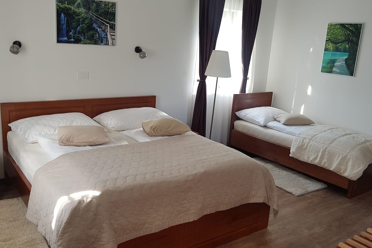 Uma cama de casal com cabeceira de madeira, uma mesinha de cabeceira com abajur, e um criado-mudo com telefone e controle remoto.