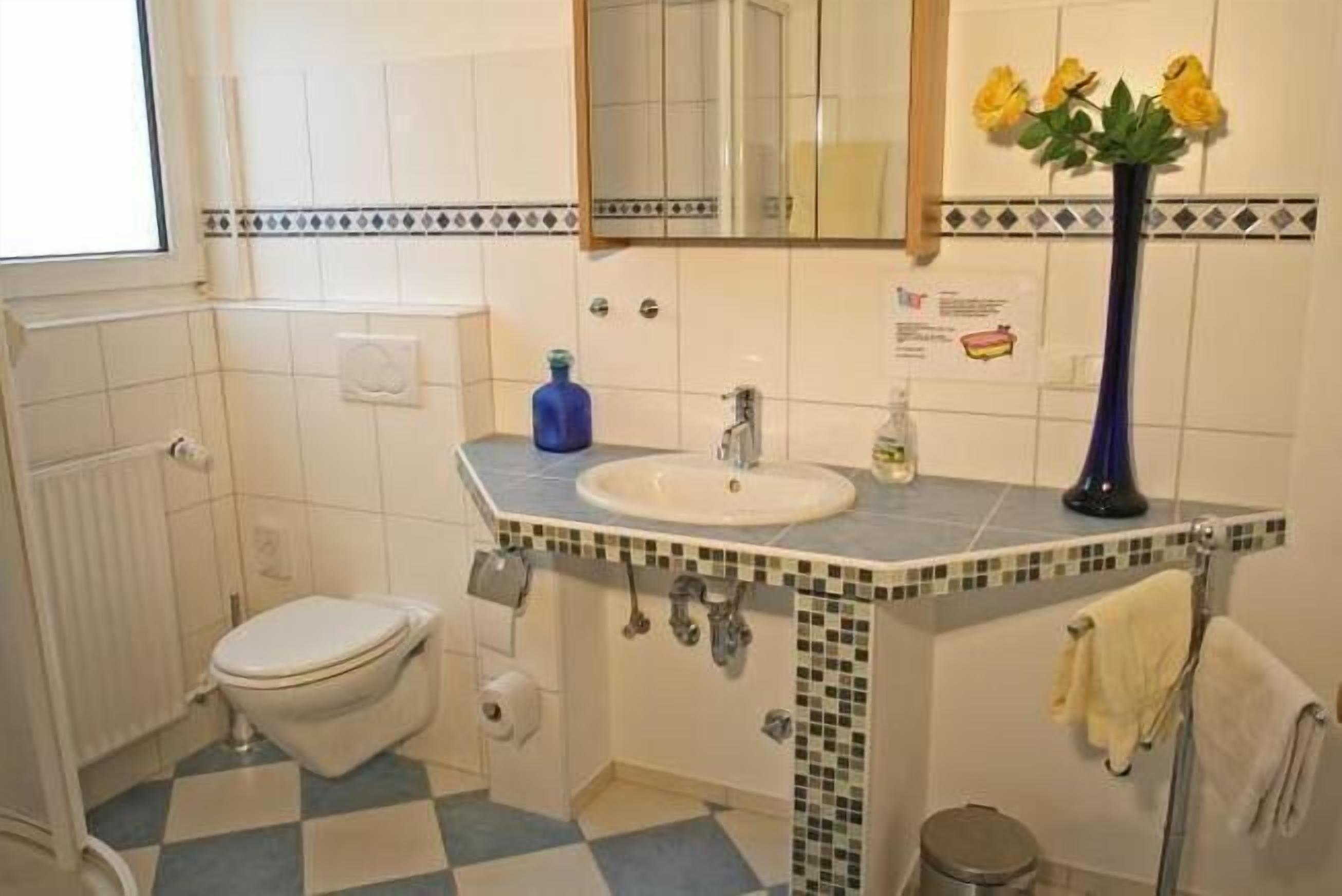 Um banheiro com vaso sanitário branco, pia com um frasco azul, espelho e um vaso com flores amarelas.